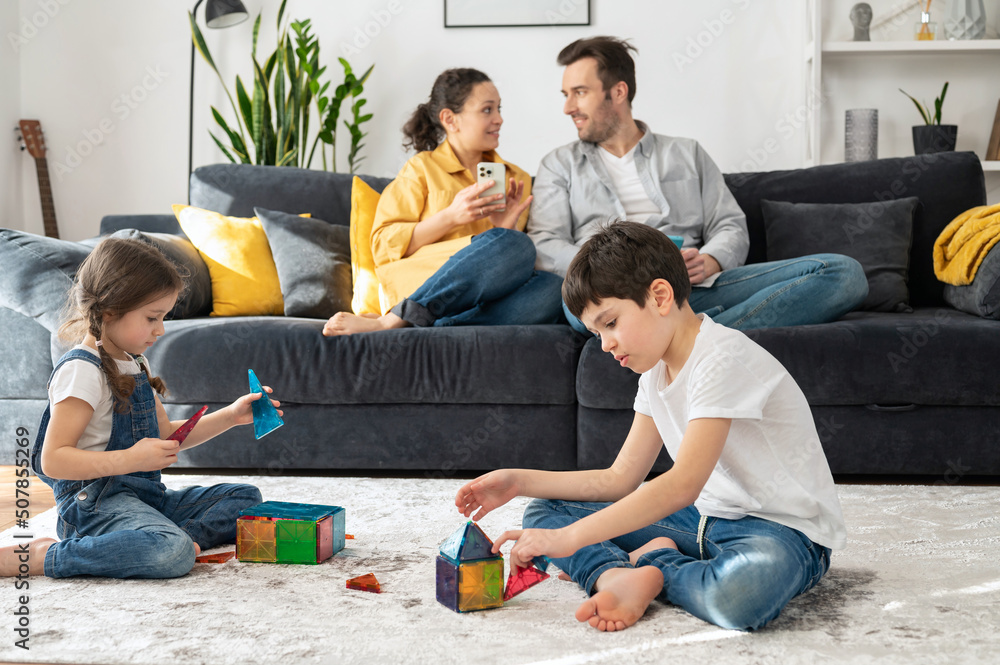 Sibling enjoying game together. Family of four at home. Two parents using smartphones sitting on ...