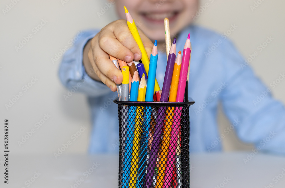 joyful kid takes a pencil from metal net box pen holder in front of him ...