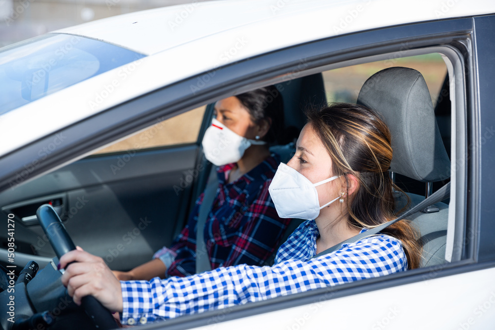 Young Hispanic woman in medical face mask driving car with female ...