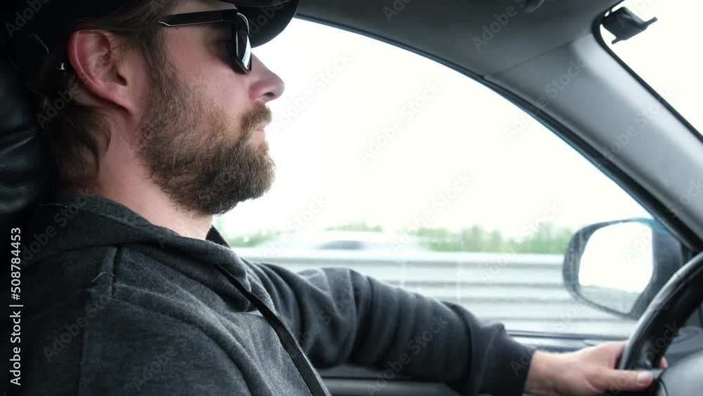 Bearded man in a cap and sunglasses driving a car during road trip ...