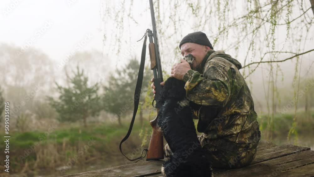 A hunter with a weapon is sitting near a misty lake next to his friend ...