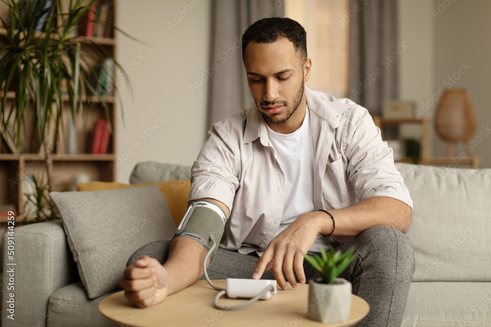 Young black man sitting on couch, checking blood pressure at home, copy ...