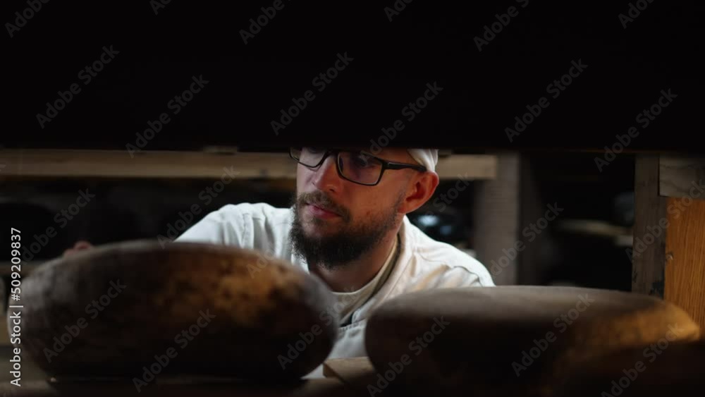 Young focused cheese maker examining cheese wheels on shelves in ...