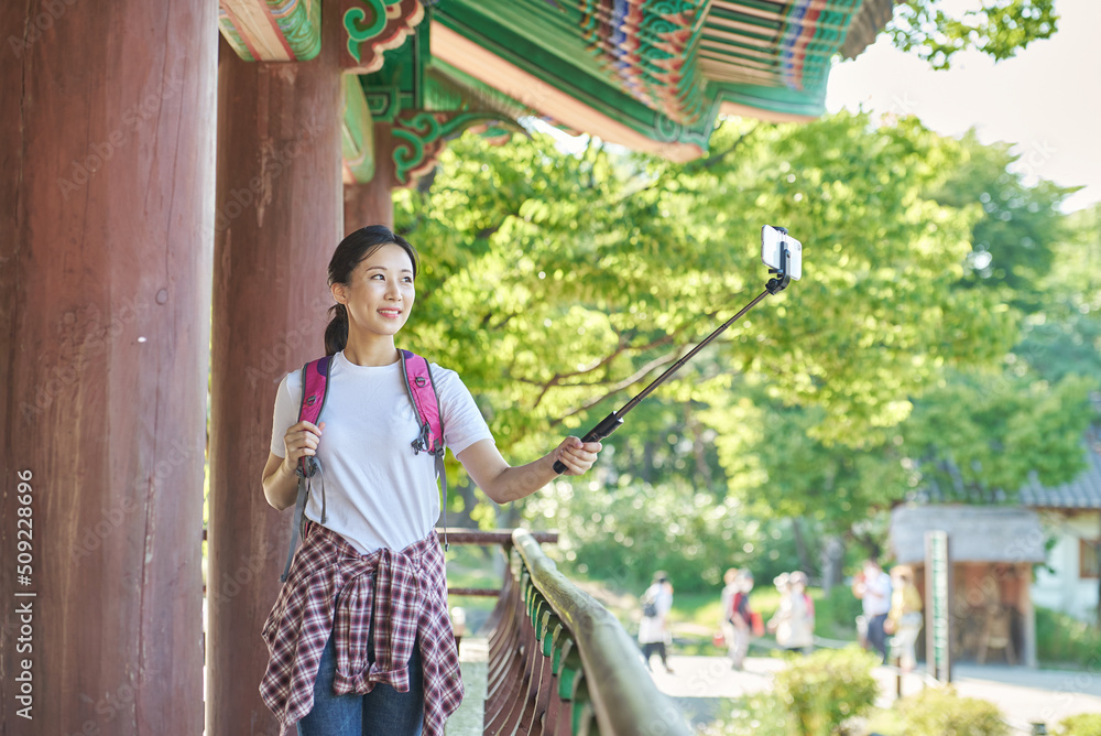 한국의 옛날 전통 가옥의 처마와 녹색 숲을 배경으로 여행하며 다양한 포즈로 스마트폰으로 셀카를 찍고있는 젊은 아시아 한국 여자