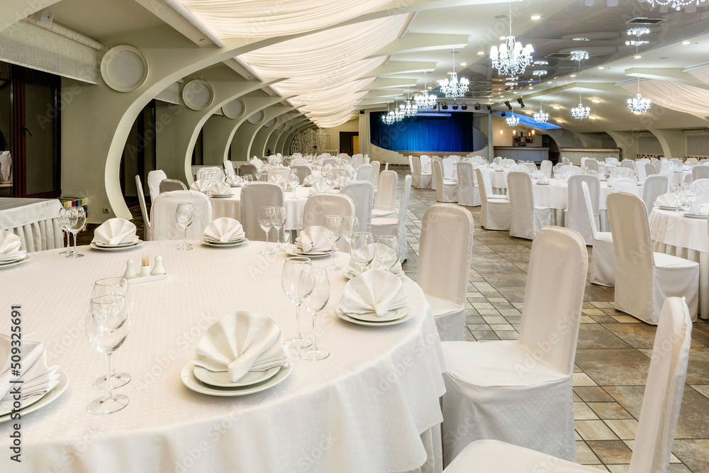 Banquet hall decorated in white with round served tables and stage for ...