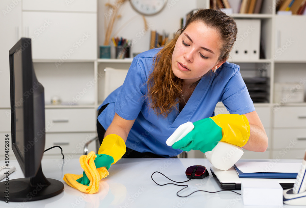 Young friendly cleaning woman in work uniform removes dust with a spray ...