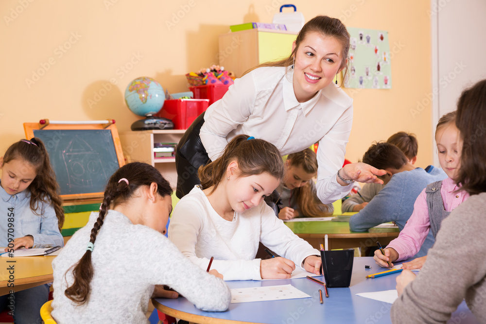 Smiling boys and girls sitting and teacher helping drawing in classroom ...