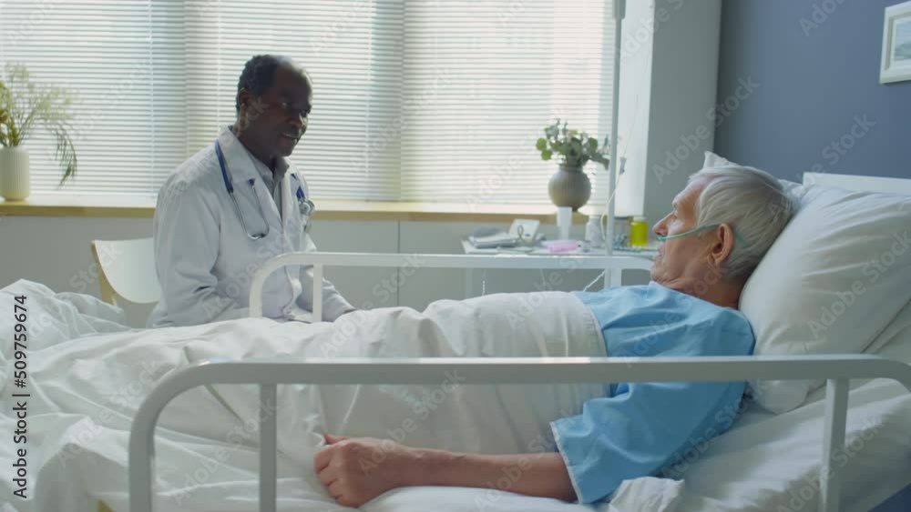Zoom in shot of African American doctor sitting by bed in hospital ward ...