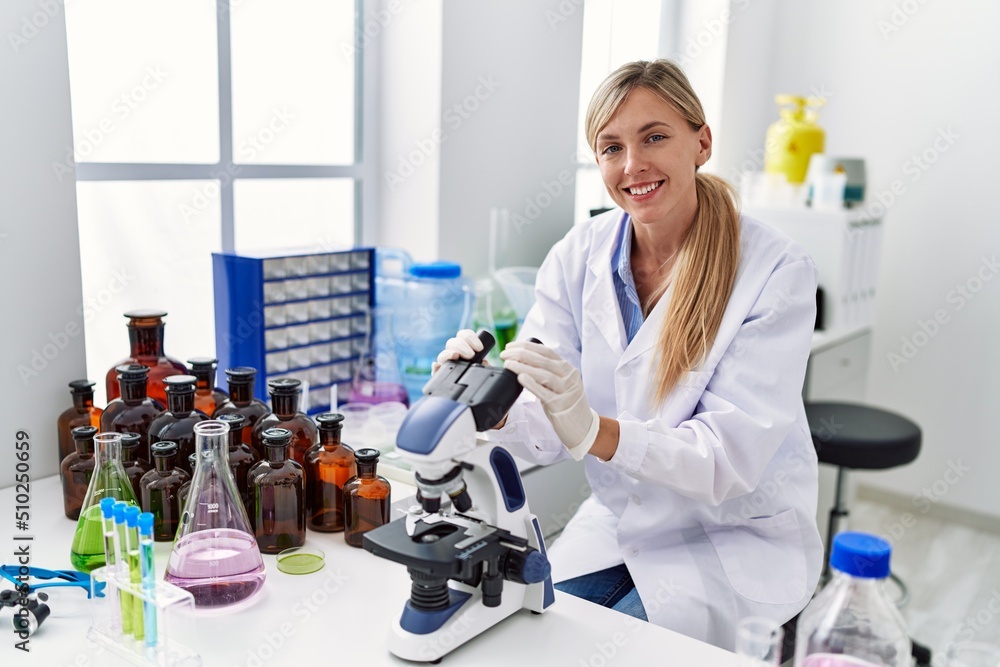 Young blonde woman wearing scientist uniform using microscope at ...