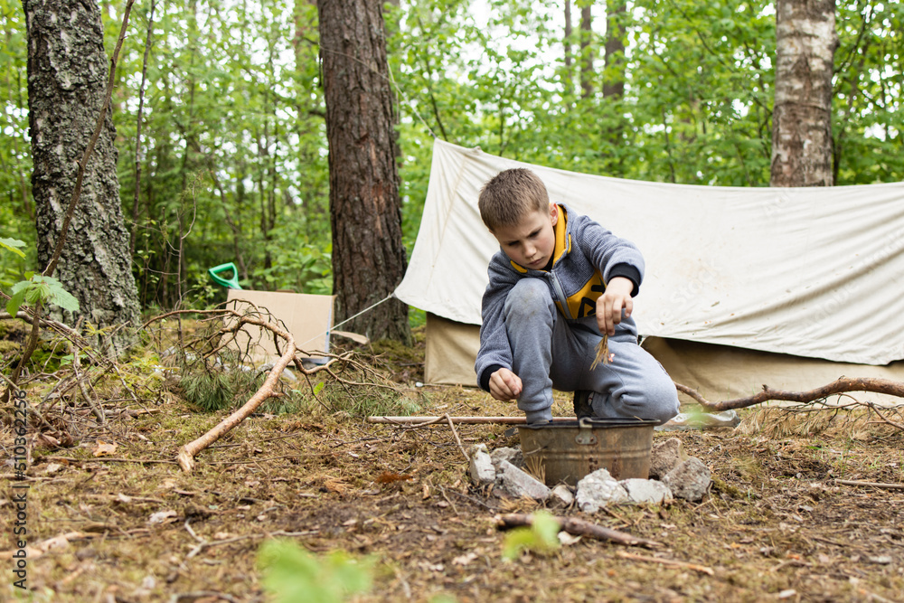 A boy building a fire in a fire pit made out of bucket near the tent in ...