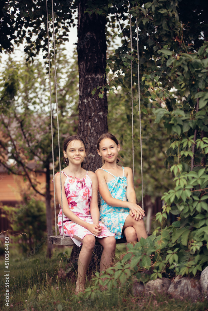 Two little barefoot girls in light summer dresses ride on an old swing ...
