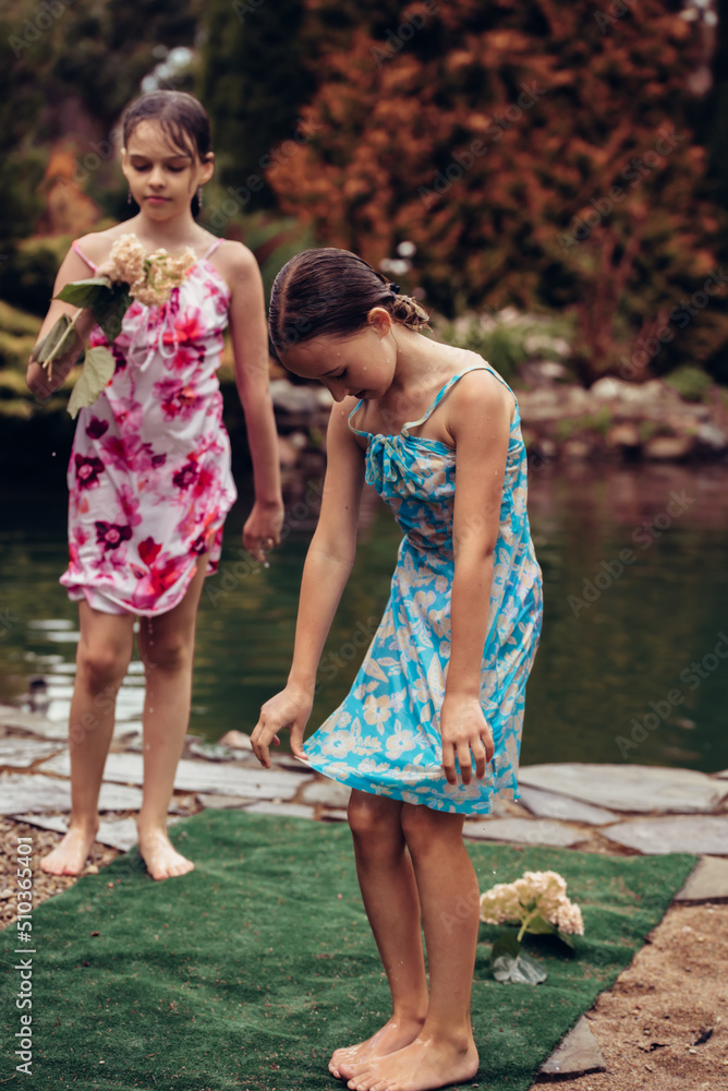 Two little girls in light summer dresses pose by the pond with flowers ...