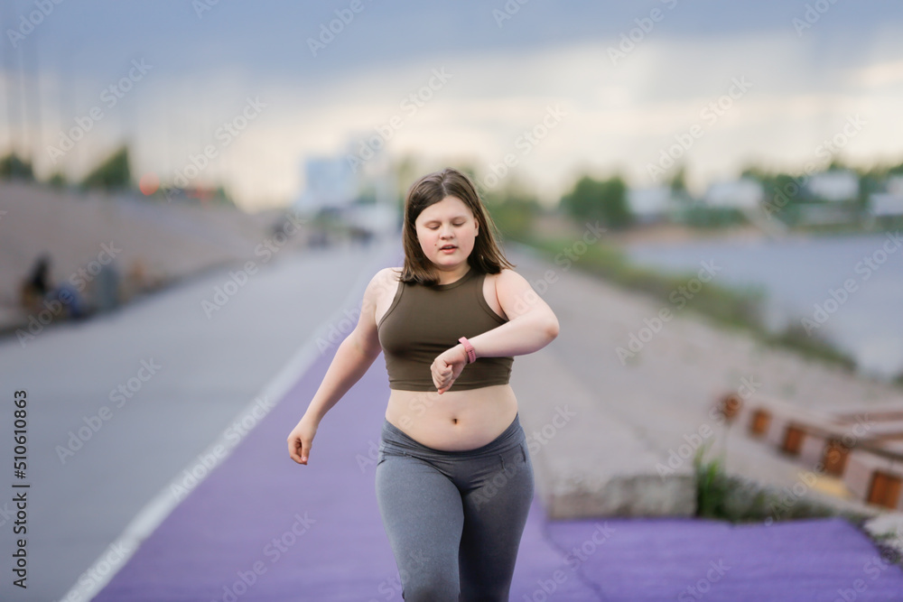 European teenage girl overweight on jog on treadmill along embankment ...