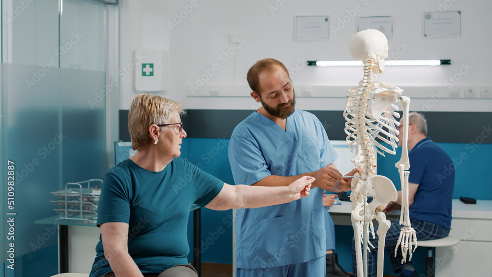 Osteopath explaining orthopedic diagnosis on human skeleton to elder ...