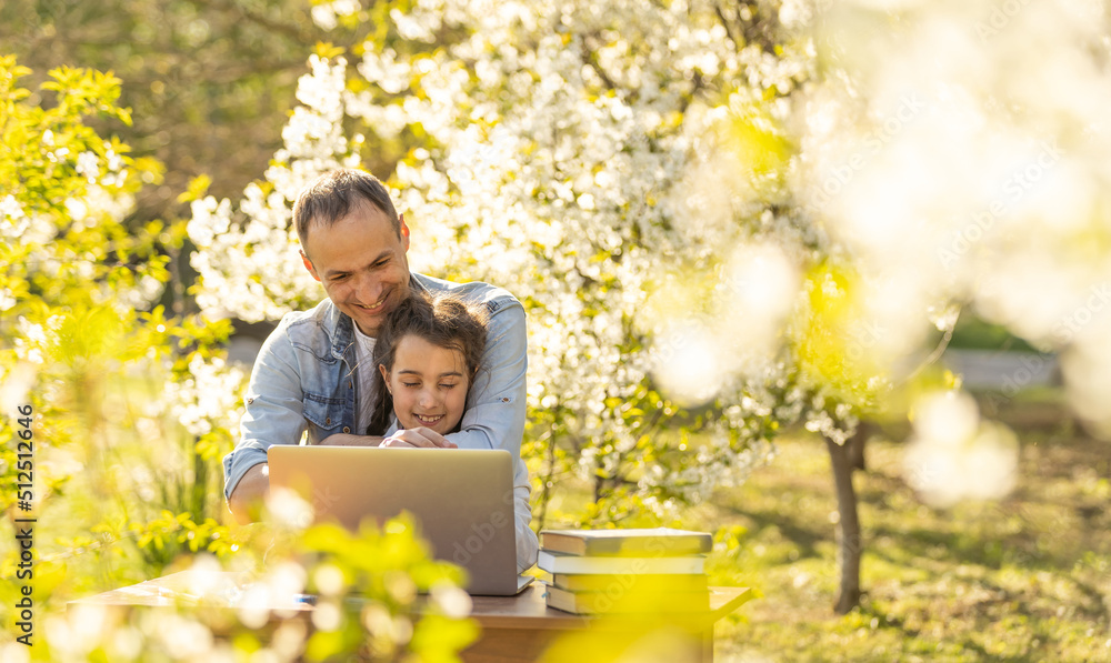 A girl and a young father are sitting at a laptop and studying in a flowered garden. Against the ...