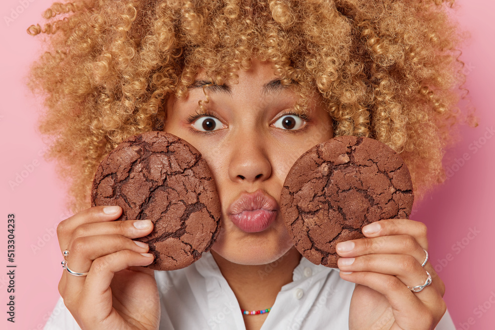 Surprised curly haired young woman holds delicious chocolate cookies ...
