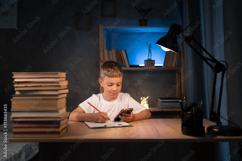 boy uses mobile phone while doing homework at night at home. kid boy ...