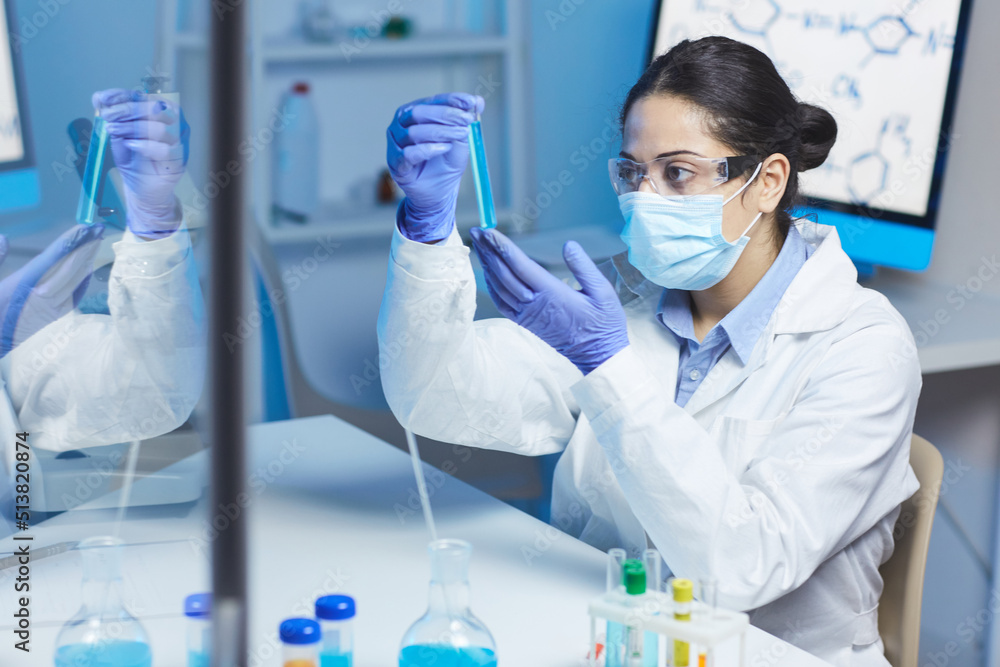 Serious young Indian female chemist in lab coat and surgical mask ...