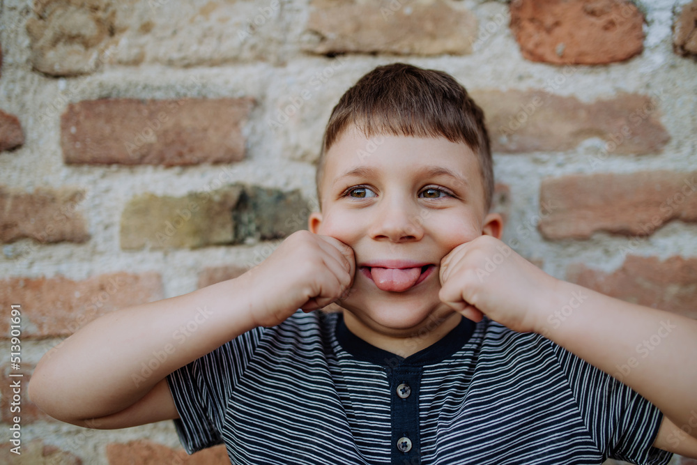 Little boy standing by brick wall and making funny faces in street ...