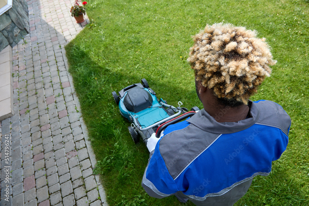 Top view of an African man in overalls mowing green grass in a modern ...