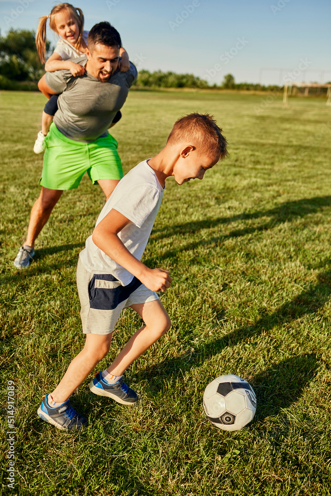Boy playing soccer by man giving piggyback ride to girl at sports field ...