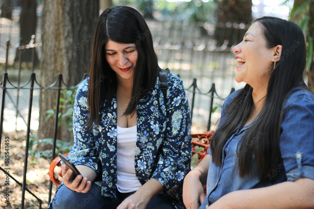Two Latin women sister friends are sitting on a park bench outdoors ...