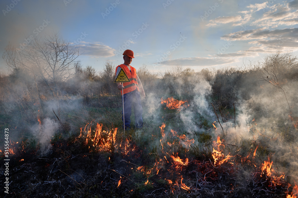 Firefighter extinguishing fire in field in evening. Man in orange vest ...