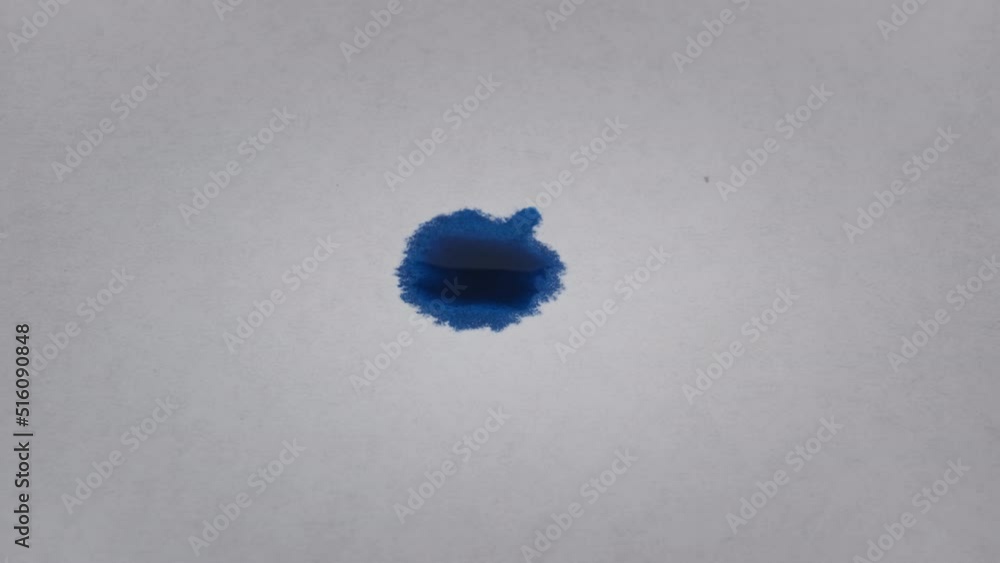 A drop of blue ink falls on white paper in macro. Ink is absorbed into ...