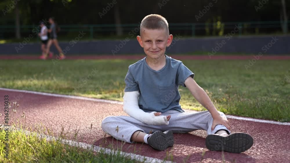 sad little boy with broken hand is sitting outdoors on sports ground on ...