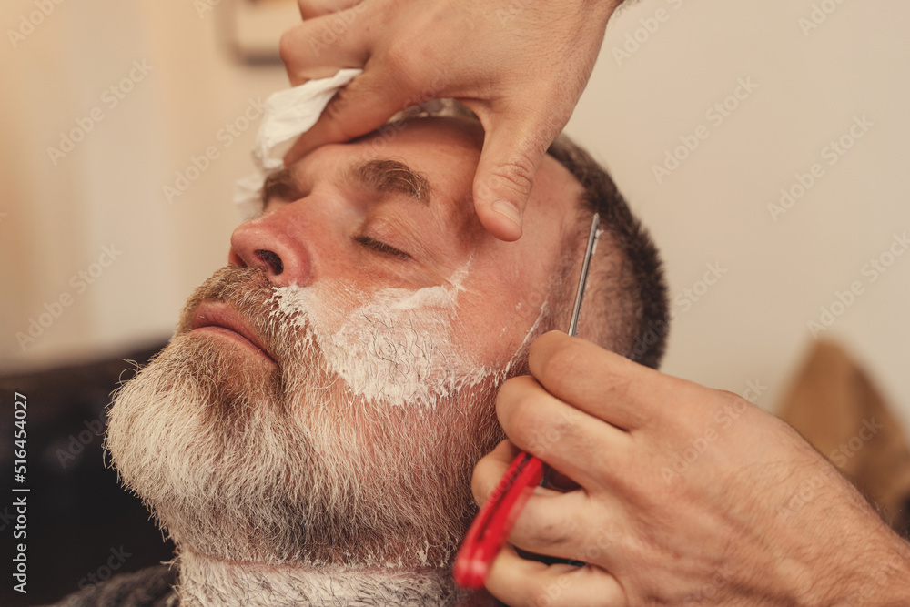 Shaving with a straight razor in a barbershop. A bearded old man being ...