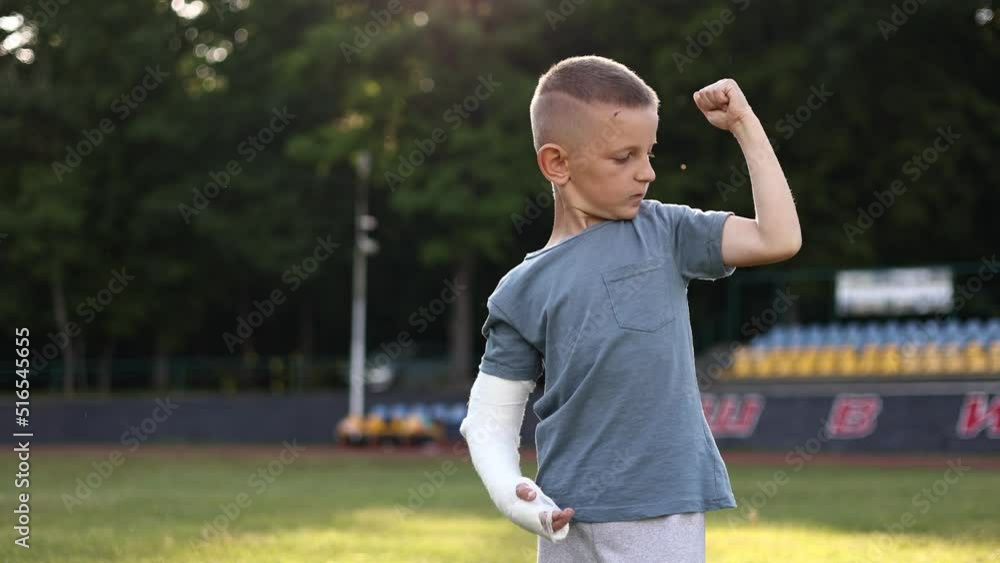smiling little boy with broken hand is sitting outdoors on sports ...
