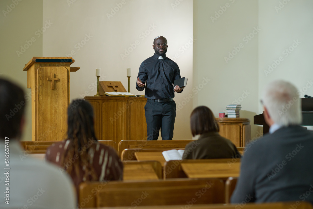 Confident priest of evangelical church with Holy Bible in hand saying sermon while standing in ...