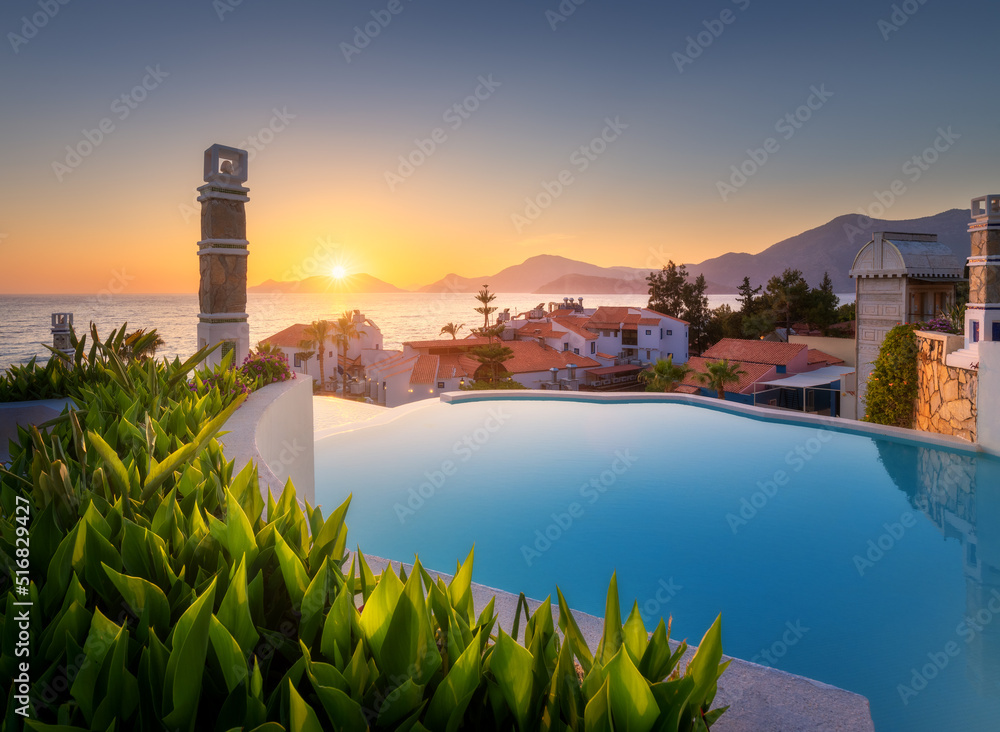 Beautiful swimming pool, green bush and orange roofs at colorful sunset ...