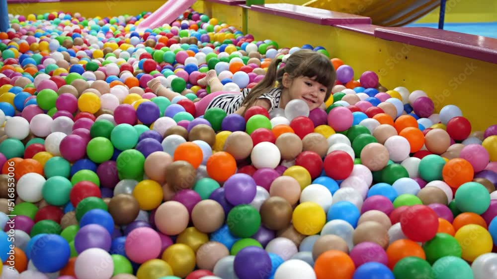 A cute little girl has fun playing lying in a pool with colorful ...