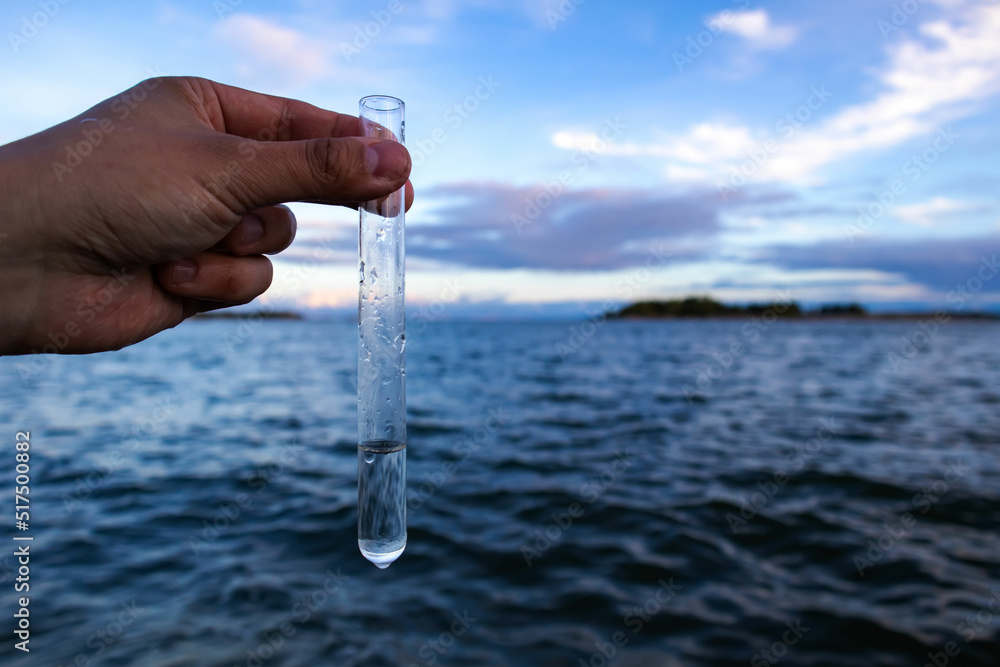 Water sample. Hand holding a test tube with water. Sea in background ...