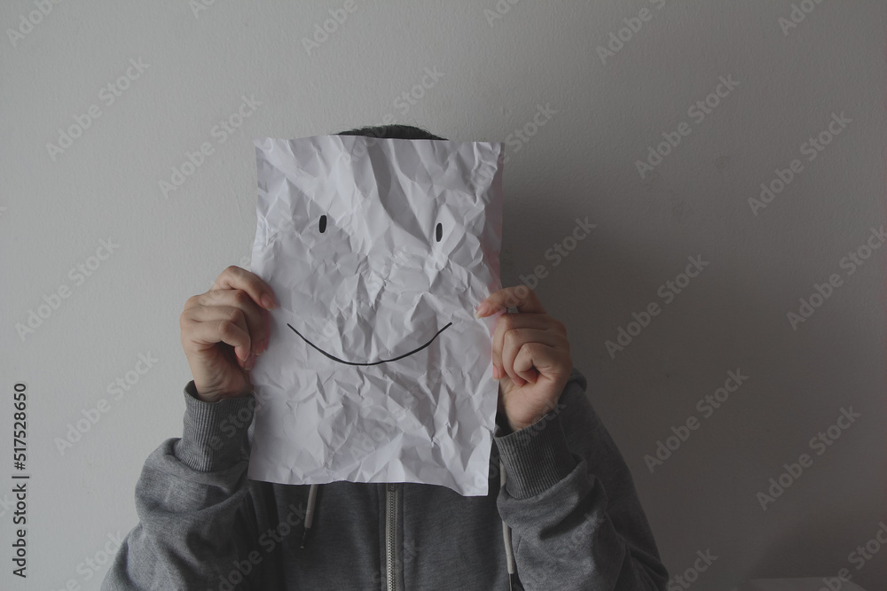 Woman holding a smiling face emoticon in front of face against a gray ...