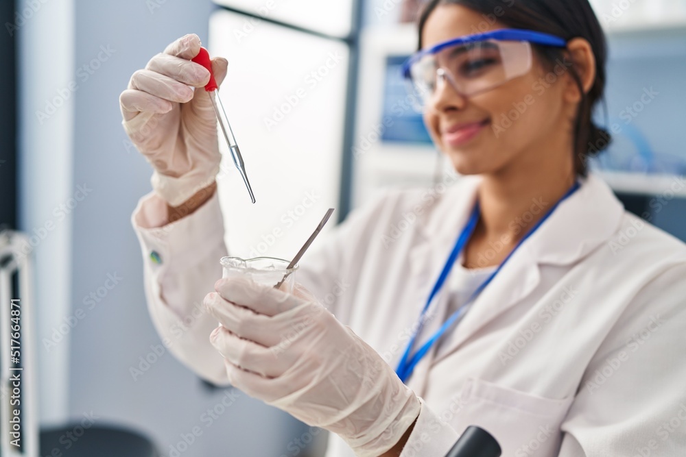 Young latin woman wearing scientist uniform using pipette at laboratory ...
