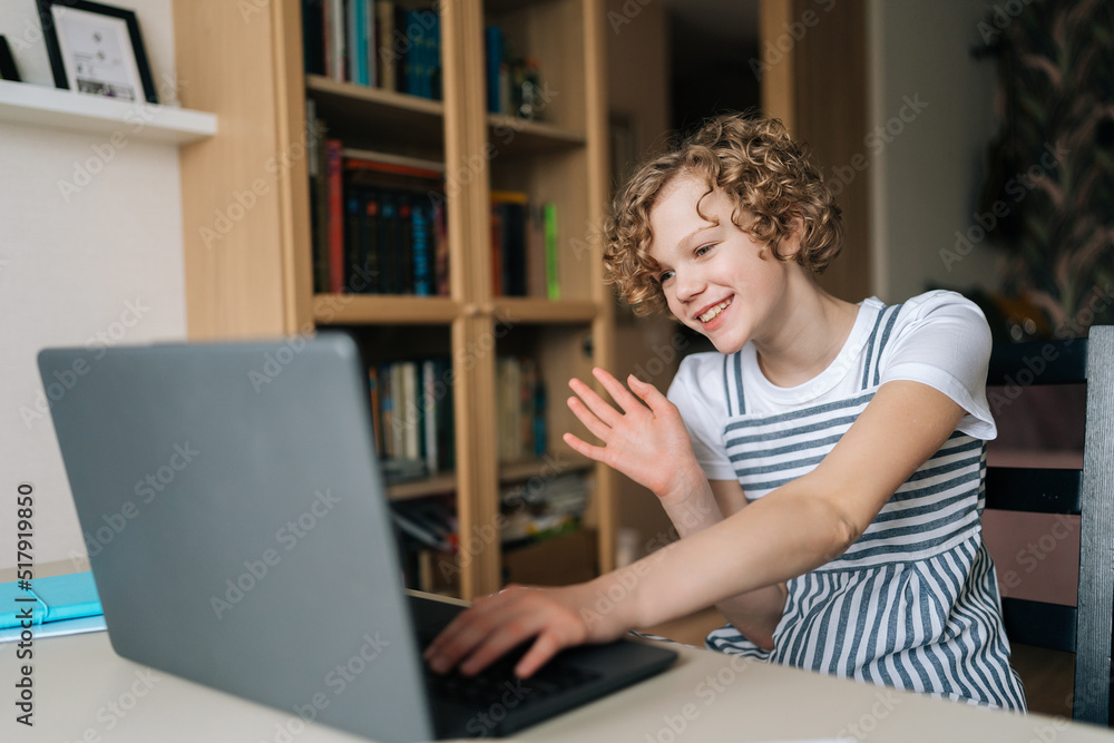 Portrait of cheerful child girl sitting at table having video call ...