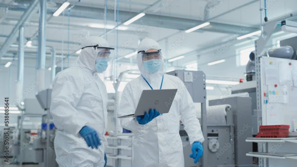 Two Scientists Standing in a Heavy Industry Factory in Sterile Coveralls and Face Masks, Using ...