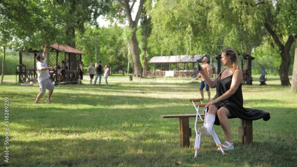 A young girl with a cast and crutches is sitting in the park, watching ...