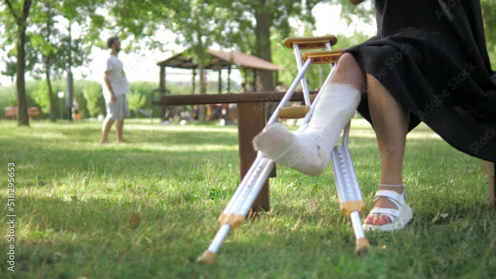 A young girl with a cast and crutches is sitting in the park, watching ...