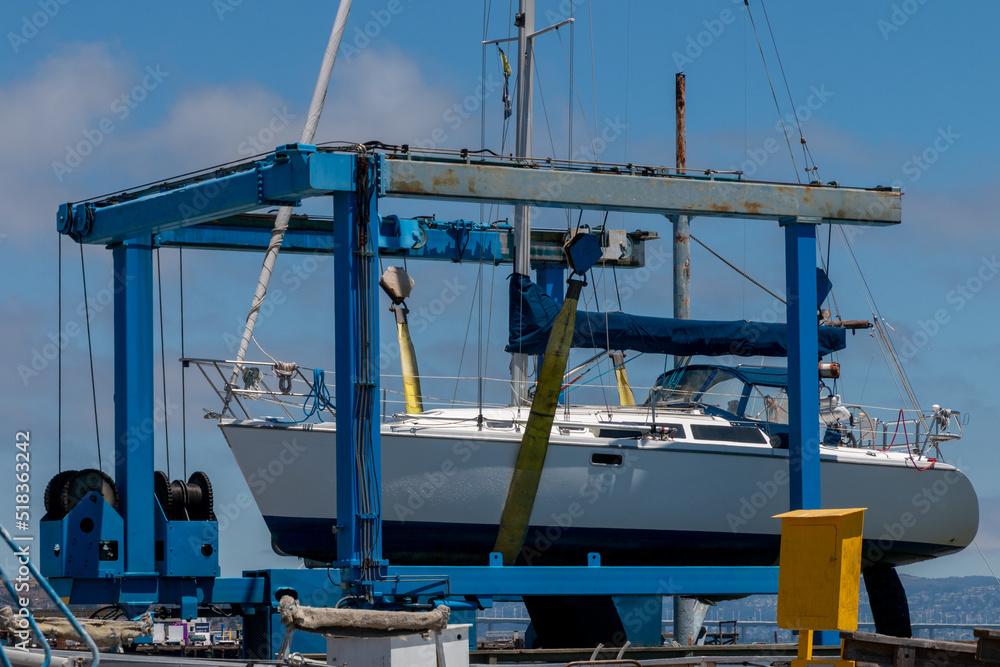 A white sail boat it being lifted out of water. The lift is blue with ...