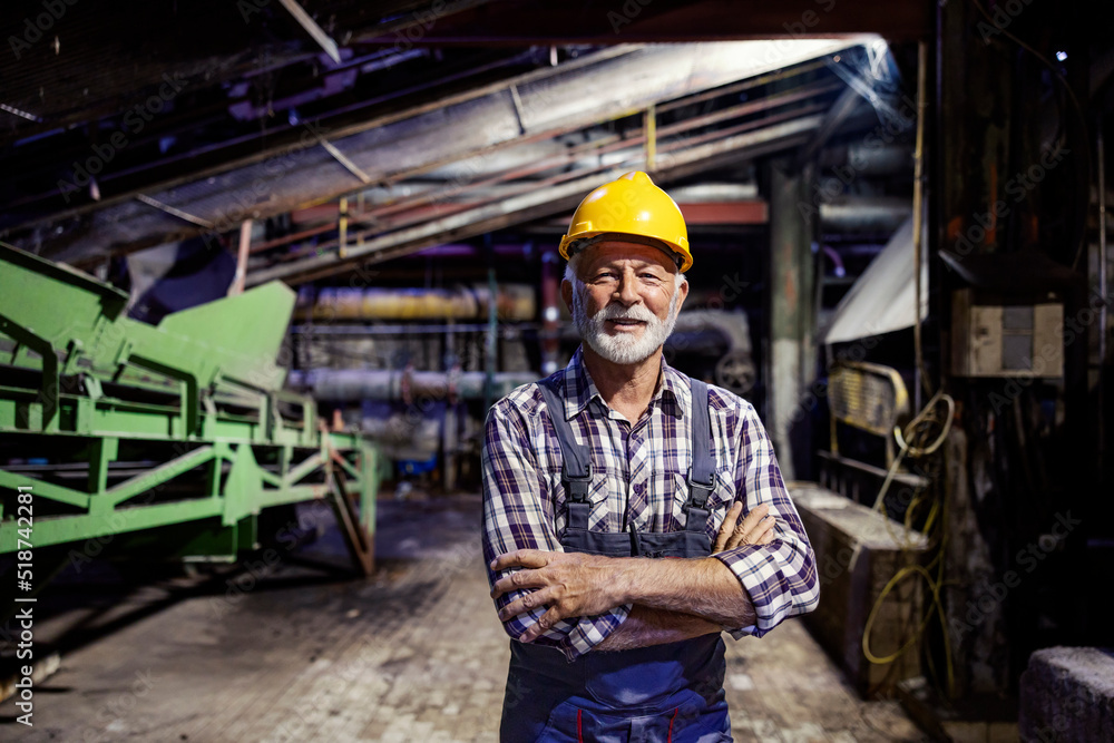 A happy senior factory worker stands in the dark with arms crossed and ...