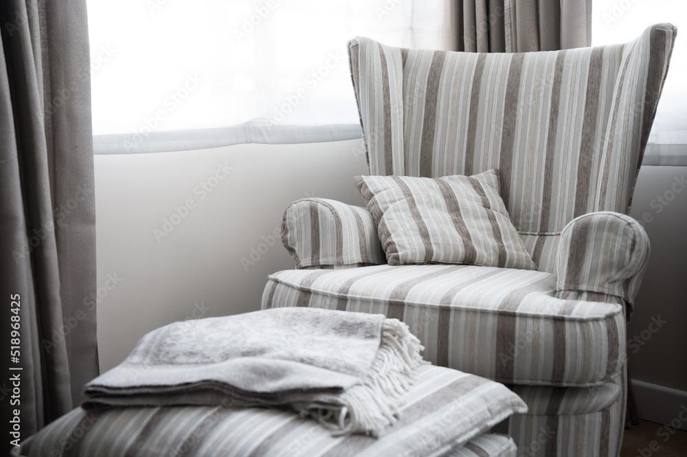 Gray wing chair, puff and blanket placed next to a window inside a ...