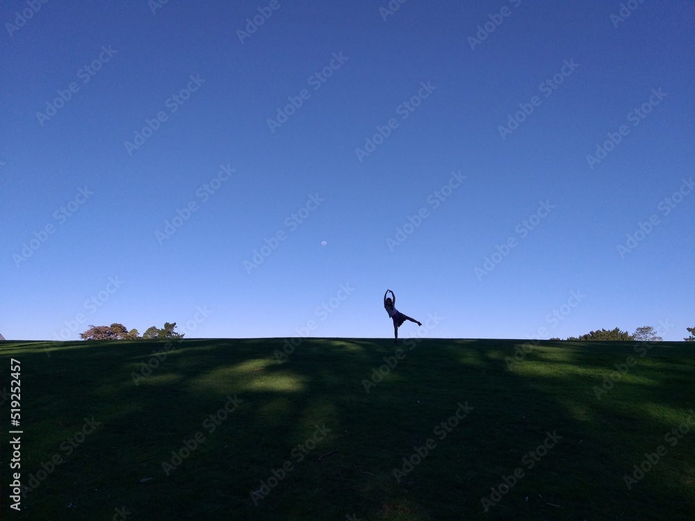Girl dance poses with her arms up high on a hill against a blue sky ...