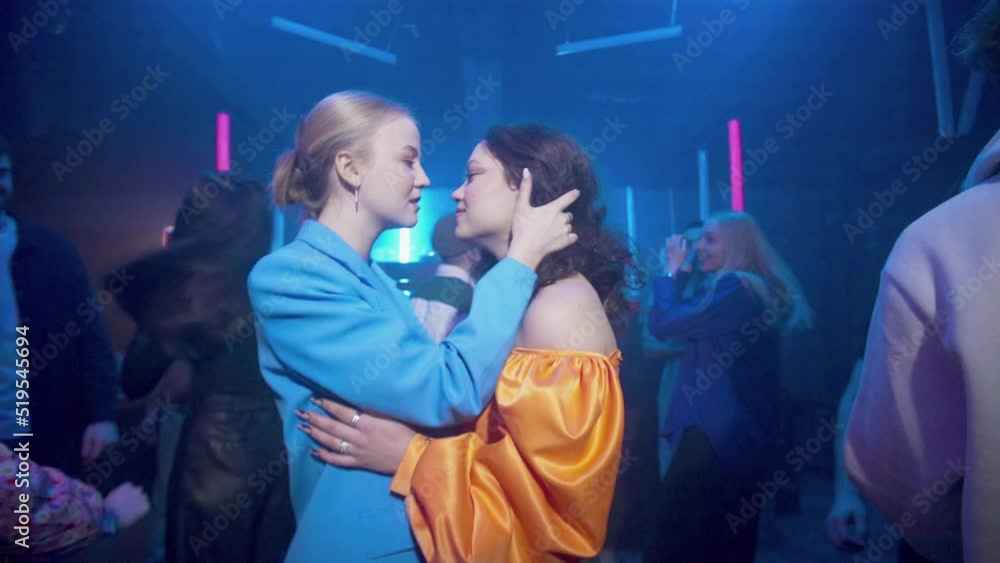 Lesbian girls approach each other and kiss among the crowd in a nightclub under the light of ...