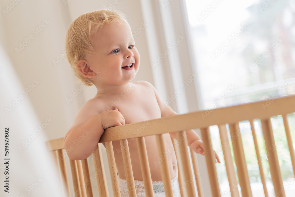 Indoor portrait of sweet little baby in diaper standing in bed trying ...
