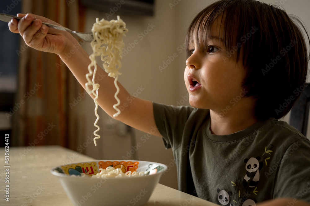 a boy with long dark hair is sitting at home in the kitchen and eating ...