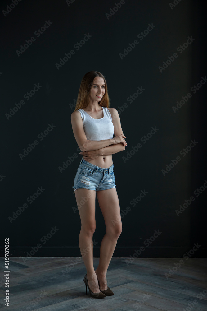 young woman in a white tank top and denim shorts on a dark background ...