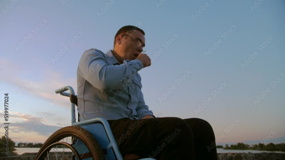man in a wheelchair and glasses is sitting and coughing.Disabled young ...
